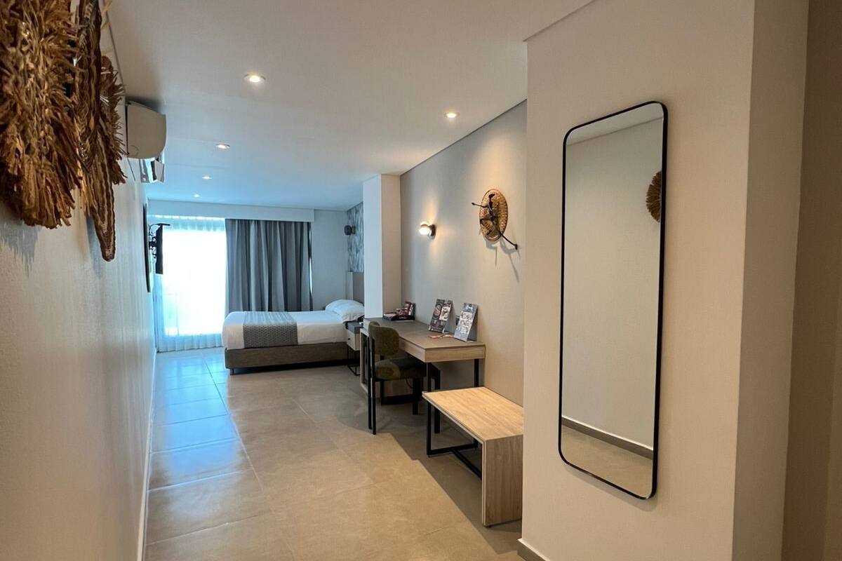 Quarto de hotel moderno com uma cama grande, uma escrivaninha, uma cadeira, um espelho e um painel decorativo na parede.