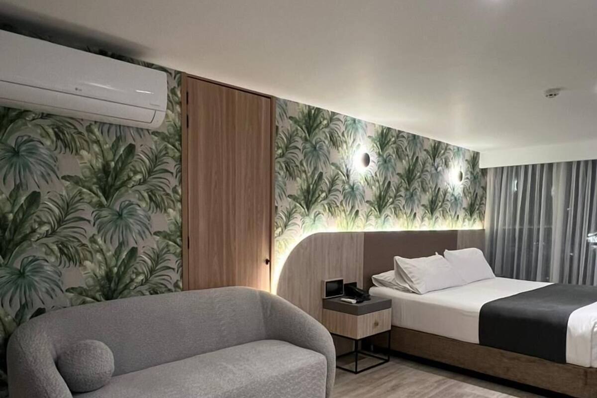 Quarto de hotel moderno com sofá, cama, mesa de cabeceira e parede com design de papel de parede tropical.