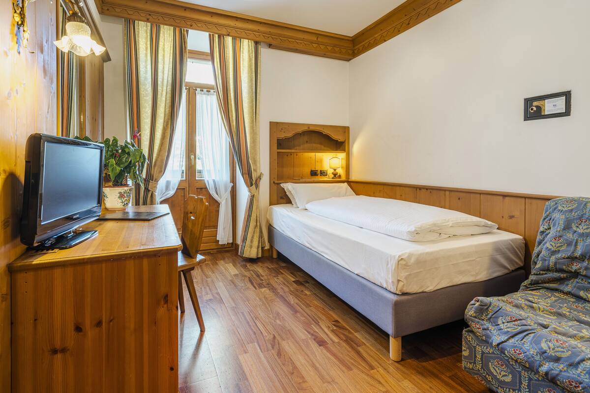 Quarto com cama, escrivaninha com televisão, cadeira, piso de madeira e janela com cortinas.