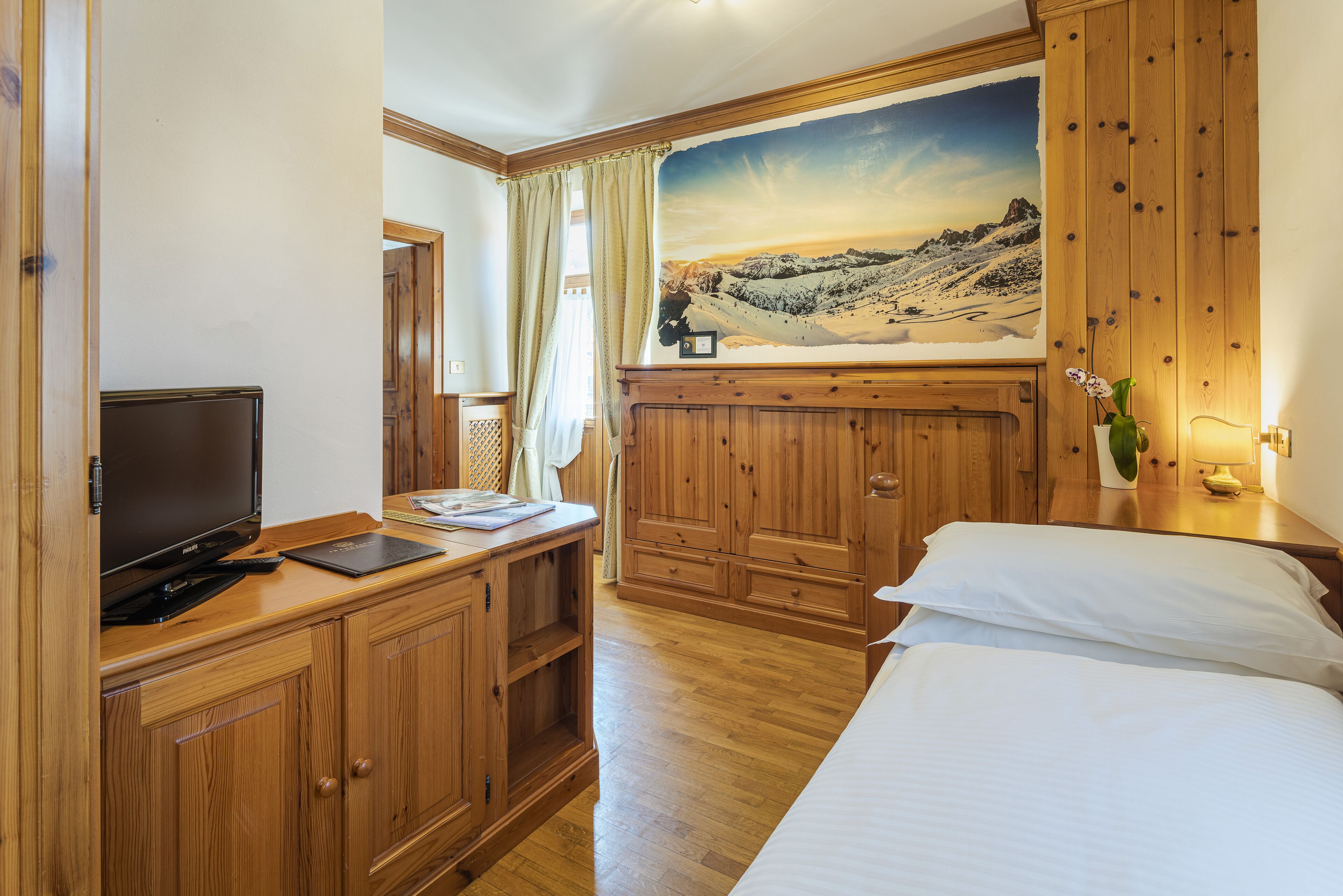 Quarto de hotel com móveis de madeira, um quadro de uma paisagem nevada, uma cama com roupa de cama branca e uma escrivaninha com televisão e comando remoto.