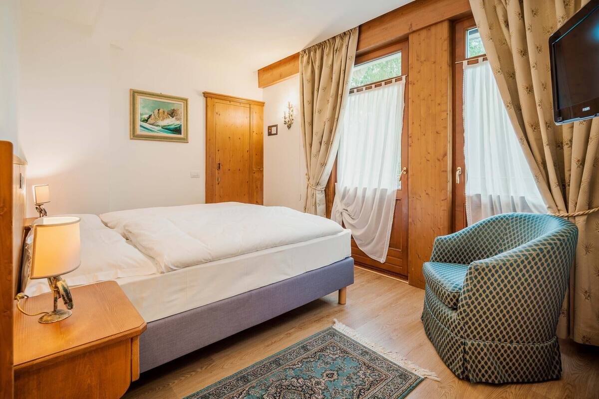 Um quarto com cama, uma cadeira, mesa de cabeceira com abajur, guarda-roupa de madeira e um quadro na parede.