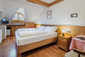 Quarto com cama de madeira, escrivaninha, cadeira, mesinha de cabeceira com abajur e janela com vista para árvores.