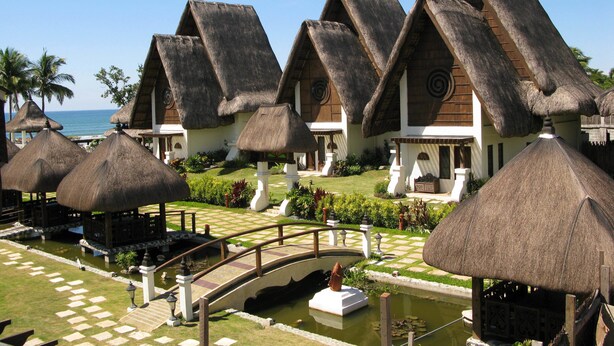 Resort com chalés de telhado de palha, uma ponte e uma piscina.