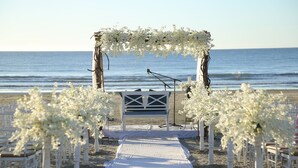 Decoração de casamento na praia, com um corredor branco, cadeiras e um suporte de microfone.