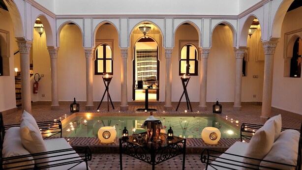 Uma área de piscina interna luxuosa, com uma mesa central, duas poltronas de descanso e uma fonte decorativa.