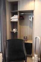 Quarto de hotel com guarda-roupa de madeira, uma cadeira estofada preta, um ventilador branco e uma mesinha com telefone e um quadro emoldurado.