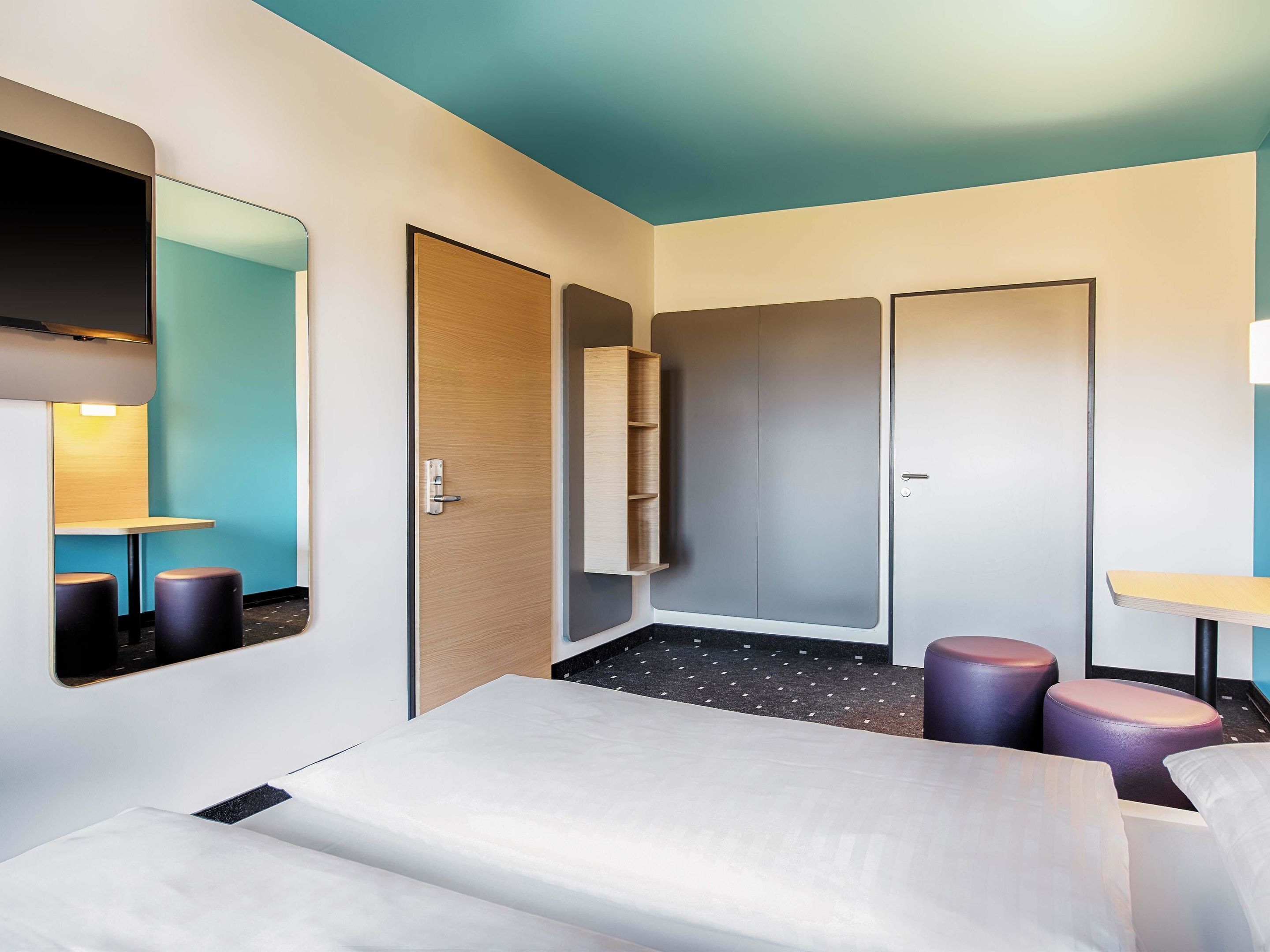 Quarto de hotel com cama, espelho, guarda-roupa e uma pequena área de estar.