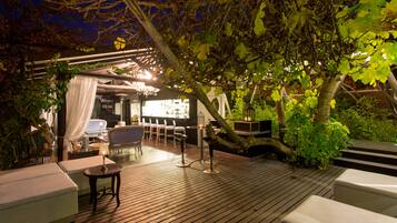 Um pátio externo com piso de madeira, assentos acolchoados brancos e um espaço de bar com adega.