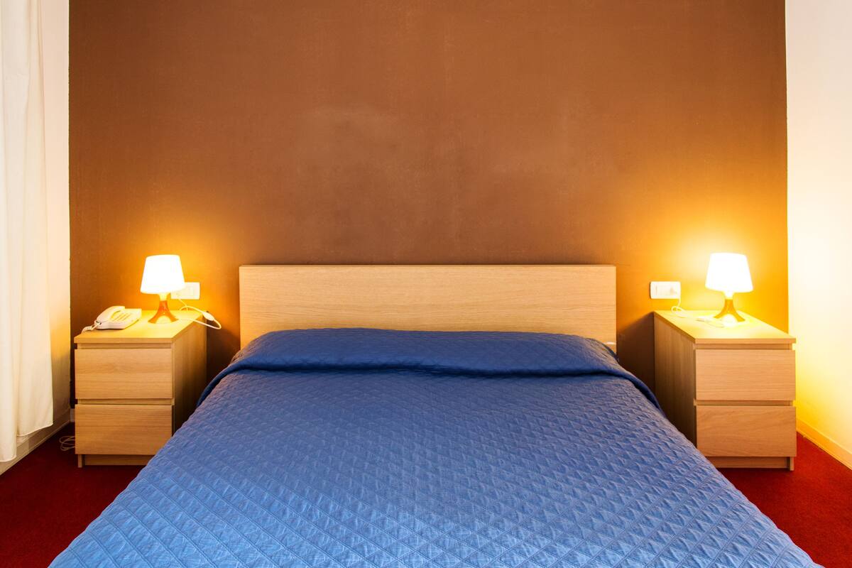 Uma cama bem arrumada com um edredom azul, duas mesas de cabeceira com abajures e um painel de cama de madeira.
