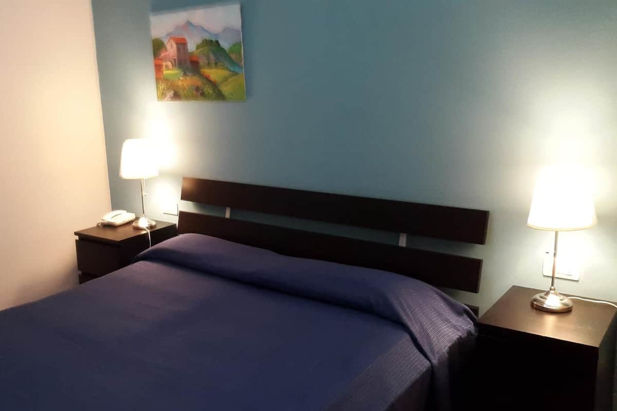 Uma cama com cabeceira, duas mesas de cabeceira com abajures, um quadro na parede e uma cortina à esquerda.
