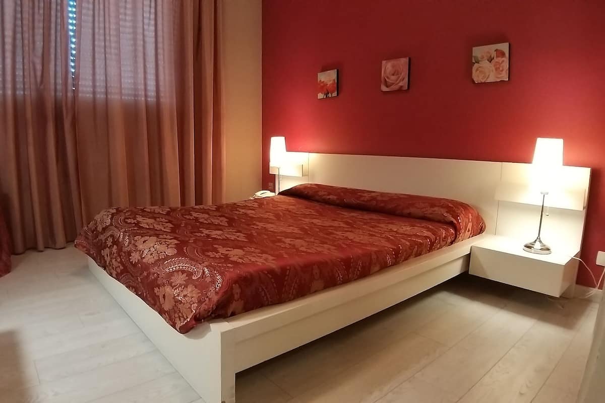 Quarto de hotel com uma cama, duas mesas de cabeceira com luminárias, uma cadeira e uma parede vermelha com três pequenas pinturas.