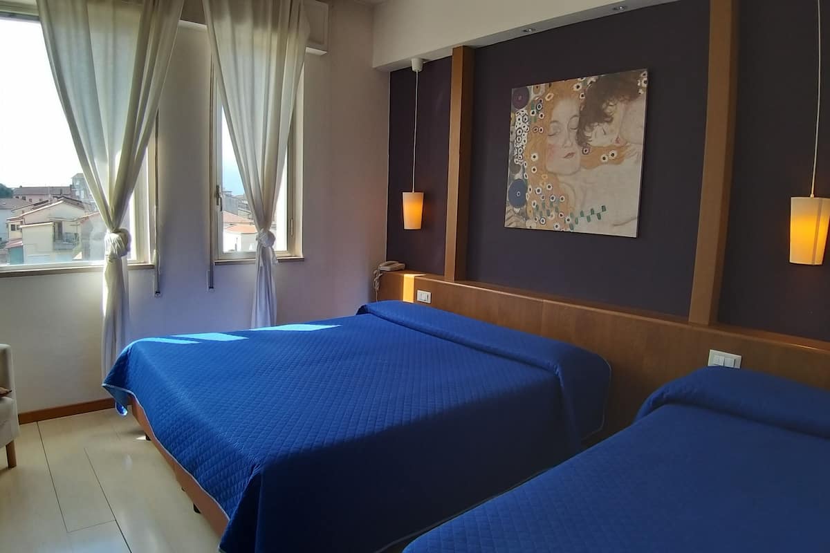 Quarto de hotel com duas camas, uma escrivaninha, uma cadeira, janela com cortinas e um quadro na parede.