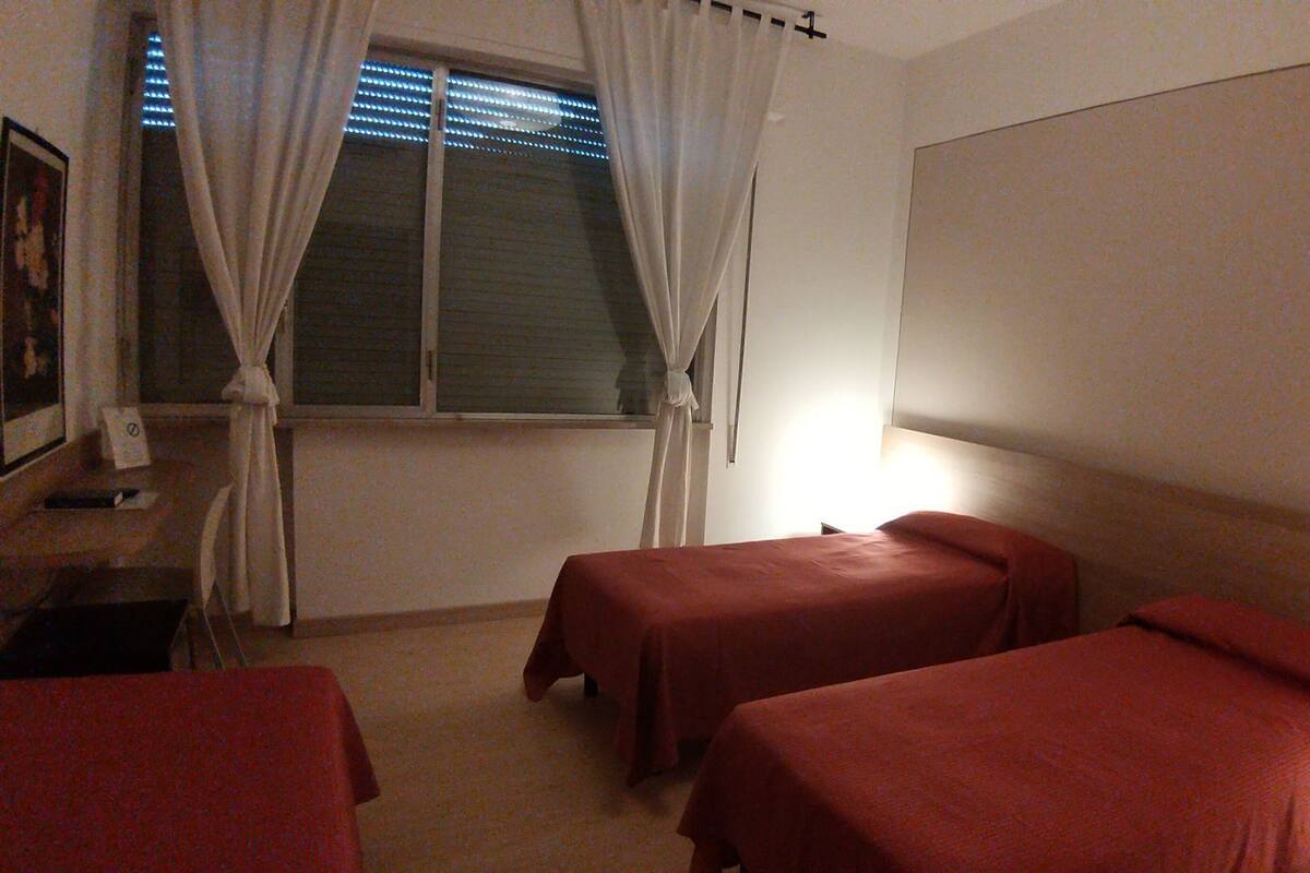 Quarto com duas camas de solteiro, uma escrivaninha com cadeira, uma janela com cortinas e uma luminária na parede.