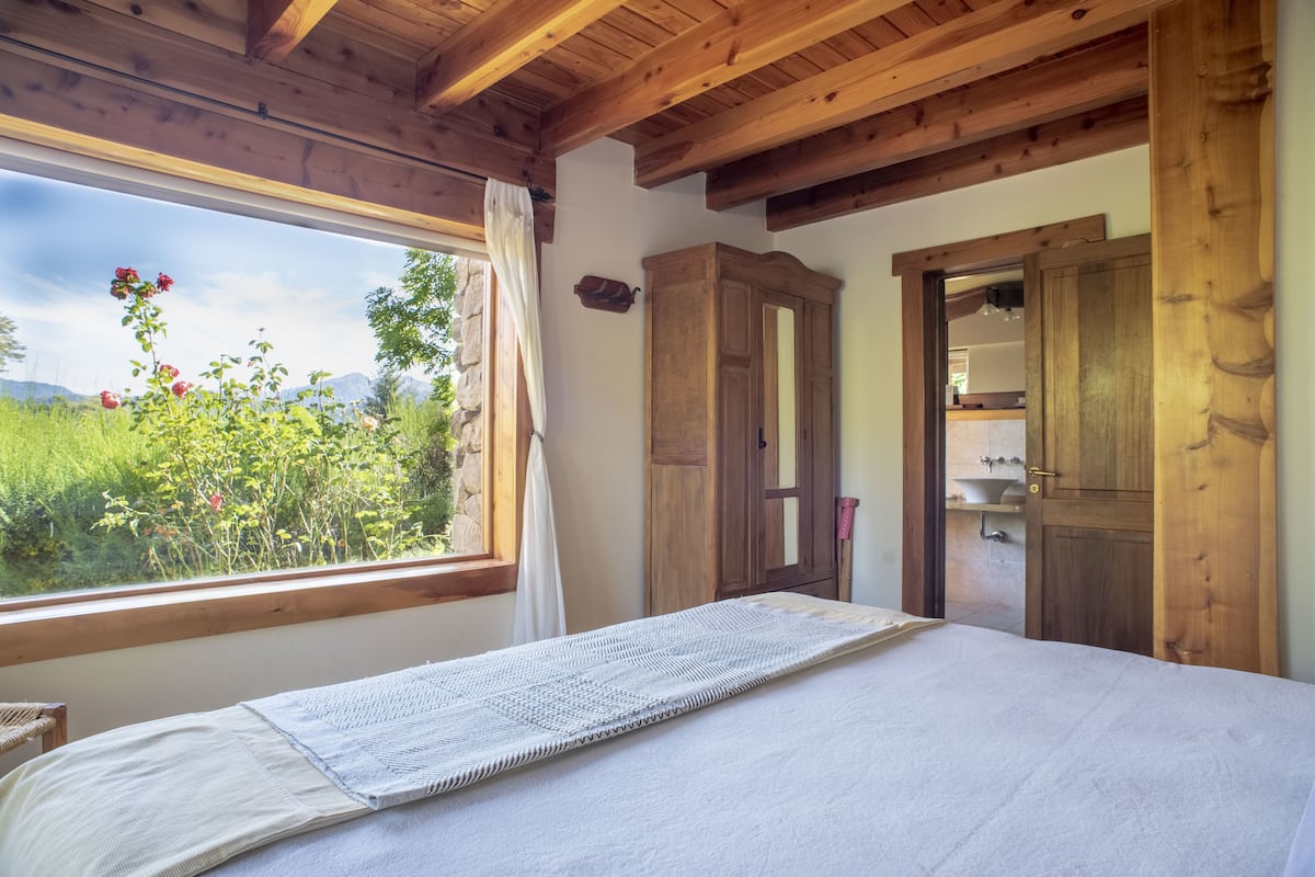 Quarto com uma grande janela, vigas de madeira no teto, cama com colchão branco, guarda-roupa de madeira e vista para a vegetação e montanhas.