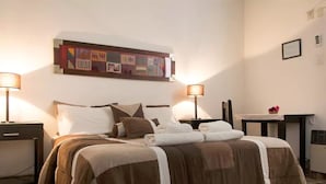 Quarto de hotel com uma cama, duas mesas de cabeceira, um criado-mudo, uma obra de arte na parede e uma pequena mesa com abajur.