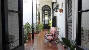 Um corredor com piso de azulejos, uma cadeira de balanço, plantas em vasos e uma porta com painel de vidro.