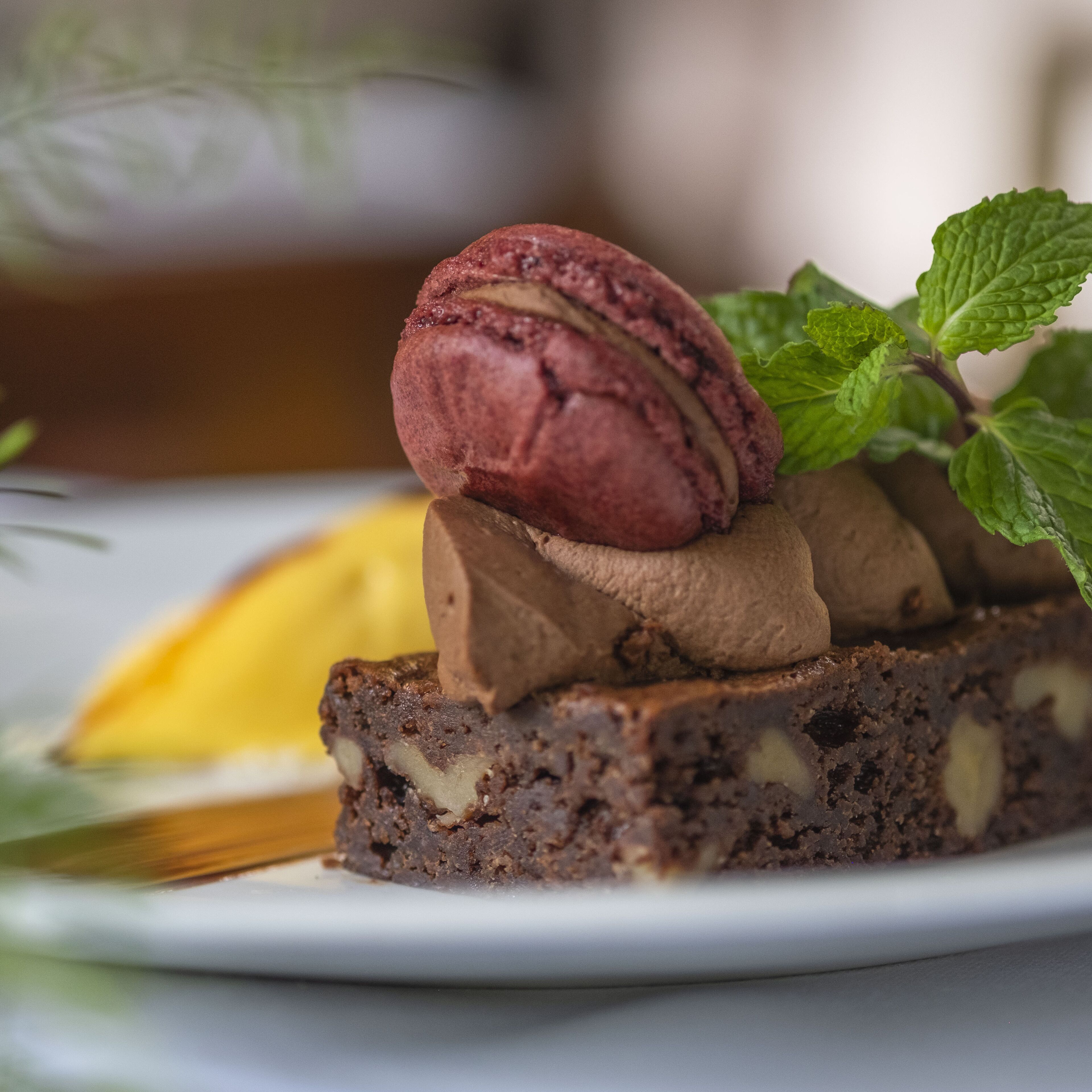 Uma sobremesa com base de brownie de chocolate, coberta com mousse de chocolate e um macaron vermelho, decorada com folhas de hortelã.