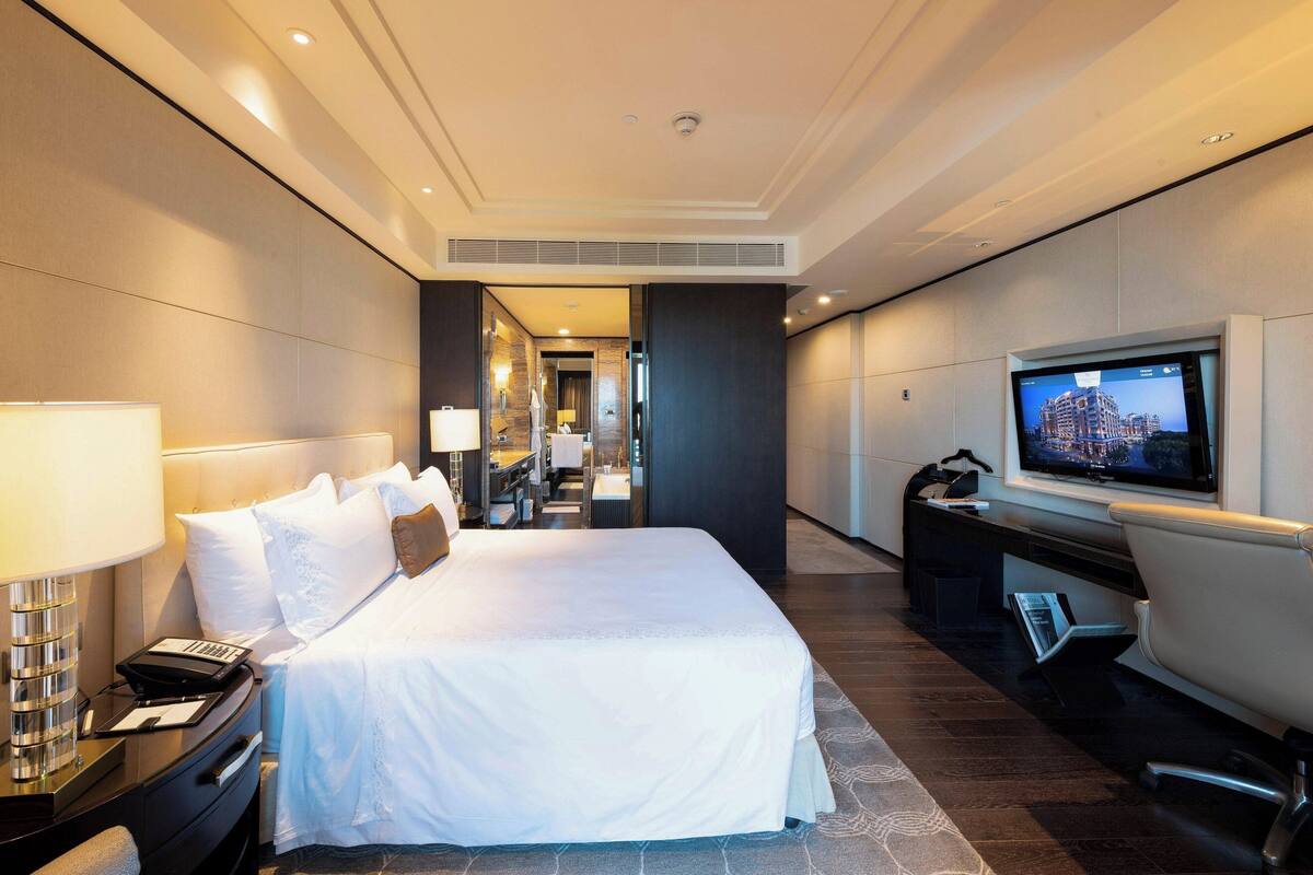 Quarto de hotel moderno com uma cama grande, uma escrivaninha com televisão de tela plana e um banheiro visível por uma porta aberta.