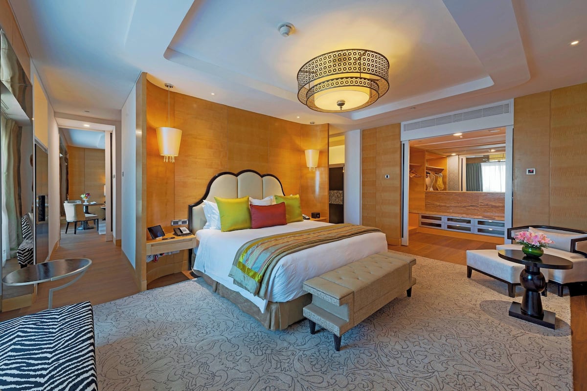 Quarto de hotel moderno com uma cama grande, mesas de cabeceira, um espaço de estar e um banheiro visível por uma porta aberta.