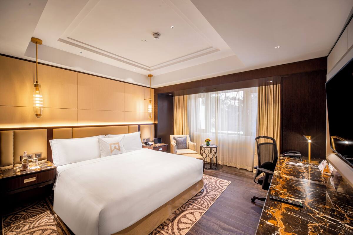 Quarto de hotel com uma cama grande, uma escrivaninha com computador, uma cadeira, uma mesinha com abajur e uma televisão.