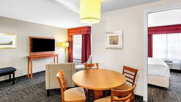 Quarto de hotel com uma mesa de madeira redonda, duas cadeiras, uma TV sobre um suporte, uma cama e uma janela grande com cortinas.