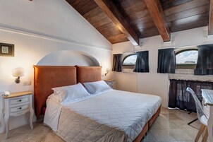 Quarto com uma cama grande, vigas de madeira no teto, mesa de cabeceira com abajur e vista pela janela com cortinas.