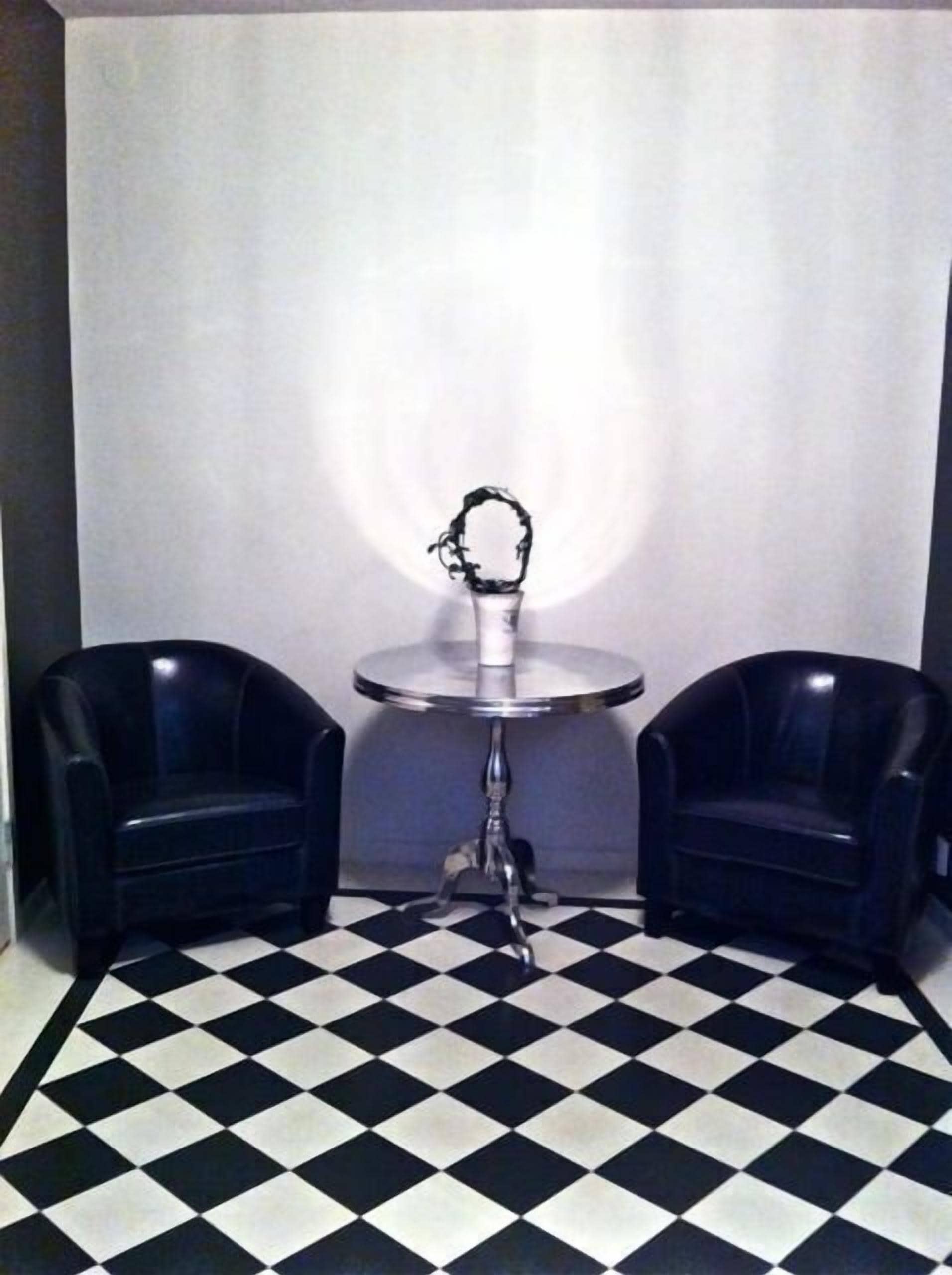 Quarto com piso xadrez, duas poltronas pretas e uma mesa redonda pequena com base metálica.