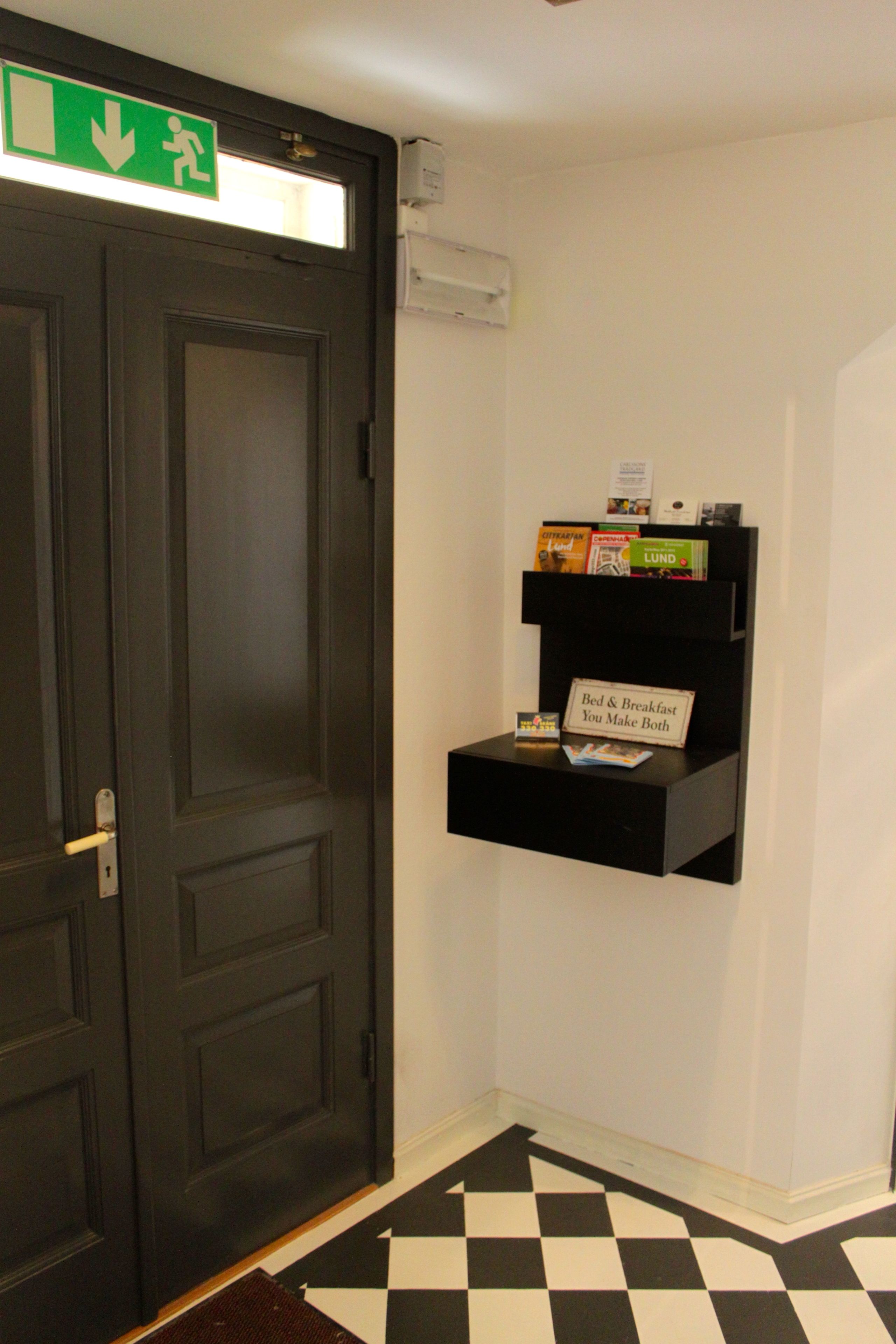 Uma porta preta com sinalização de saída de emergência, um chão quadriculado e uma pequena prateleira preta com livros e uma placa que diz "Bed & Breakfast You Make Both".