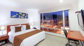 Quarto de hotel com uma cama grande, uma escrivaninha e vista para a cidade à noite.