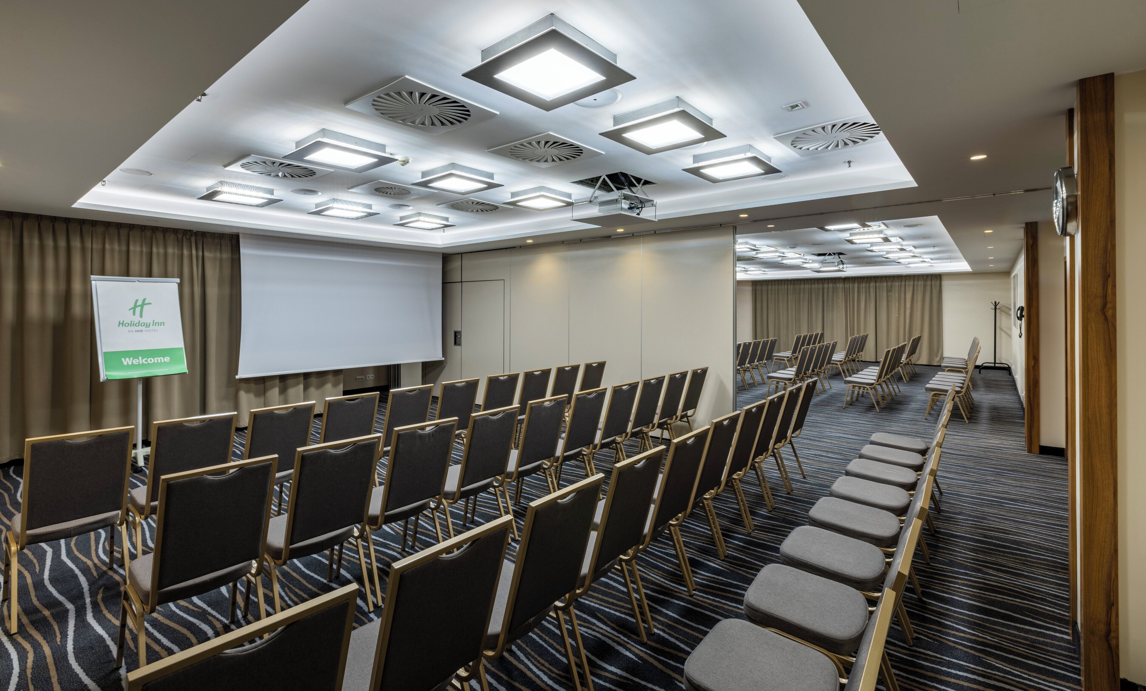 Uma sala de conferências de hotel com fileiras de cadeiras, uma tela de projeção e uma placa de boas-vindas aos hóspedes.