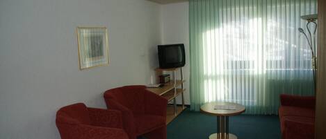 Quarto de hotel com televisão sobre um suporte de madeira, duas poltronas vermelhas, uma mesa redonda pequena e uma janela com cortinas leves.