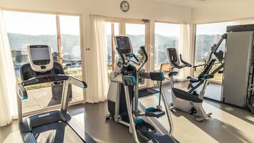 Uma academia com esteiras, elípticos e máquina de pesos, grandes janelas com vista para o exterior.