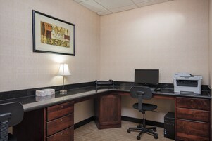 Escritório de hotel com mesa, cadeira, impressora e uma moldura na parede.