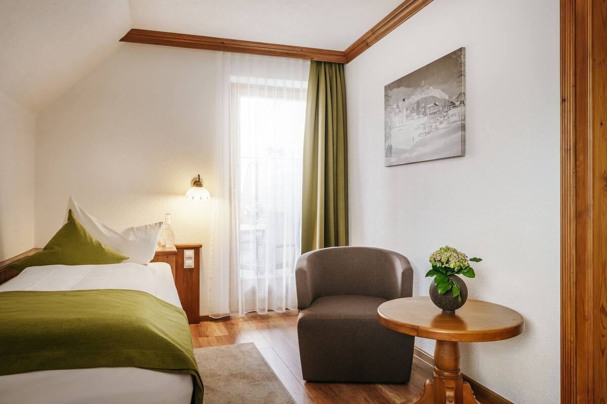 Quarto de hotel com cama, uma cadeira, uma mesinha com um vaso, uma luminária e um quadro na parede.