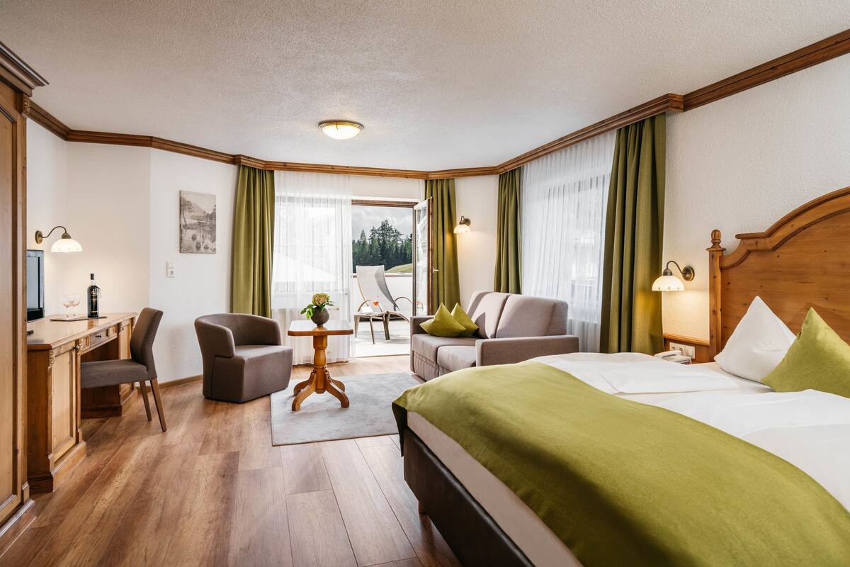 Quarto de hotel com cama, escrivaninha, cadeira, pequena mesa com planta e vista para o exterior.