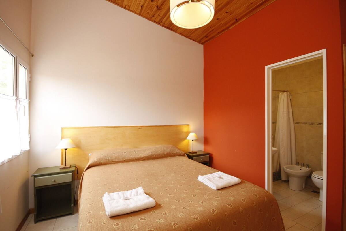 Quarto de hotel com uma cama, duas luminárias laterais, uma cabeceira de madeira, uma janela e um banheiro visível por uma porta aberta.