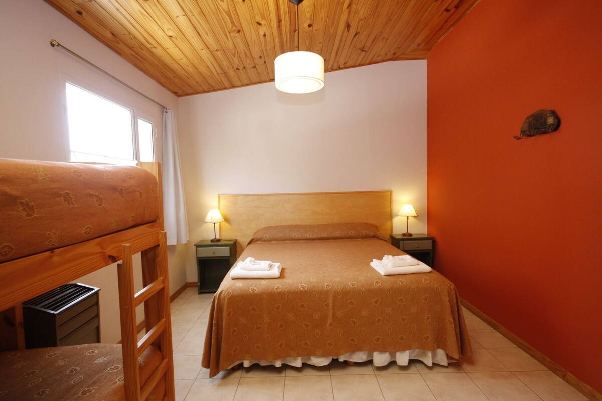 Quarto com cabeceira de madeira, duas luminárias de apoio, cama com colcha marrom e uma janela com cortinas.