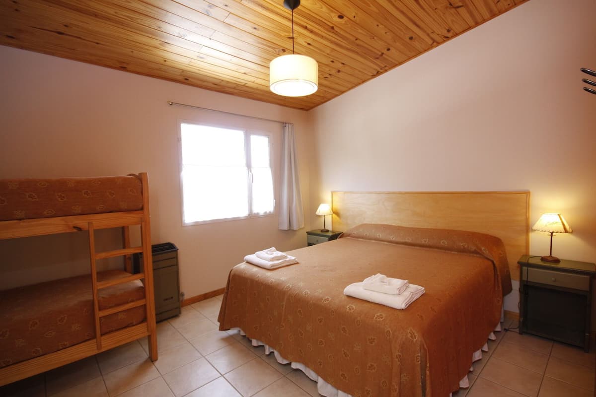 Quarto com cama de solteiro, uma cama individual, duas mesas de cabeceira com luminárias, janela com cortinas e teto de madeira.