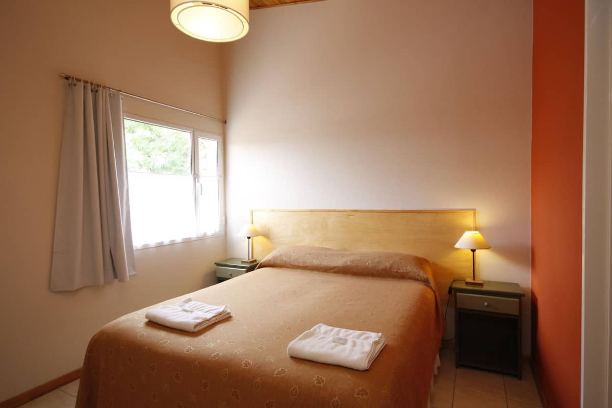 Quarto de hotel com cama, duas luminárias de cabeceira, janela com cortinas e mesa de apoio.