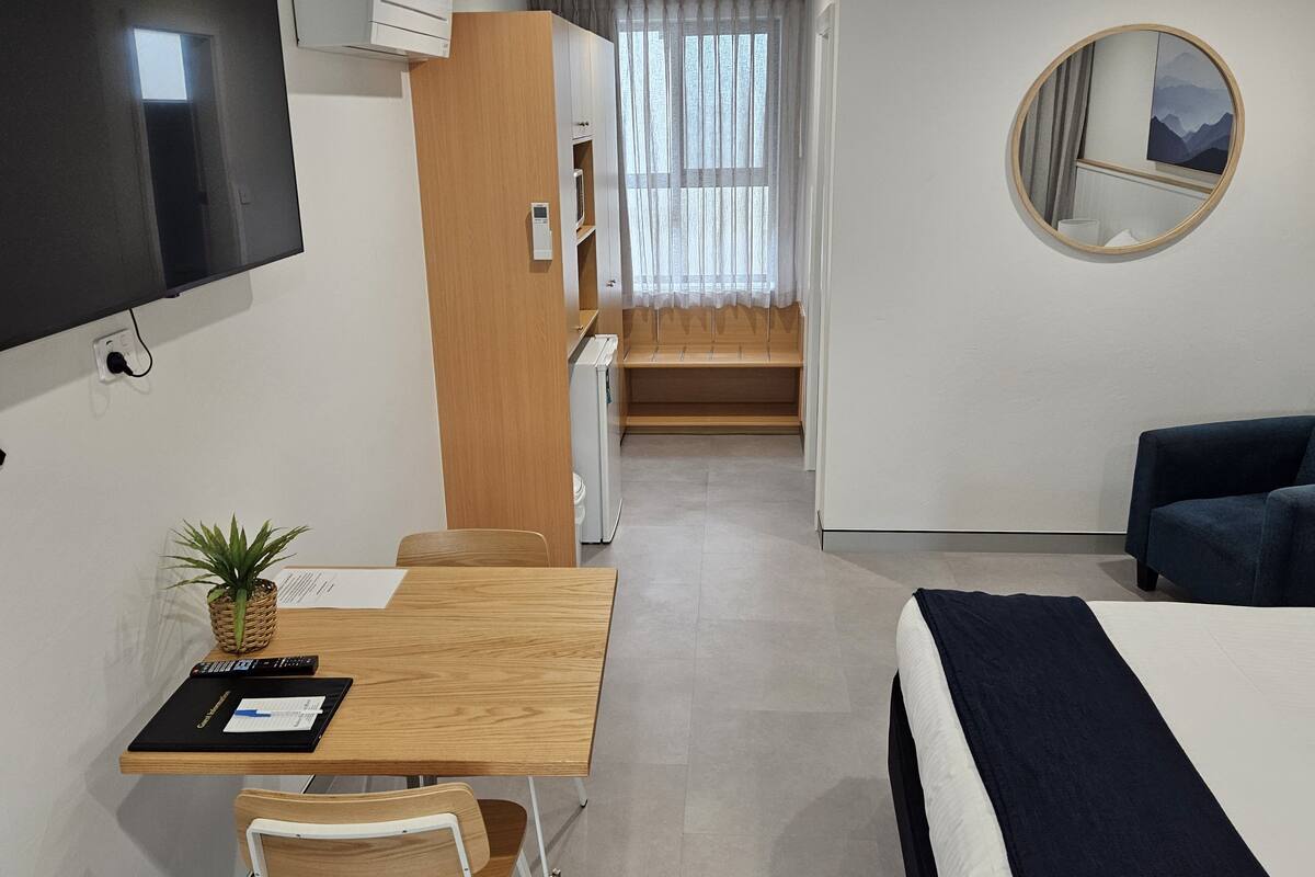 Quarto de hotel com cama, escrivaninha com cadeira, pequena mesa com planta, micro-ondas, geladeira, televisão e janela com cortinas.