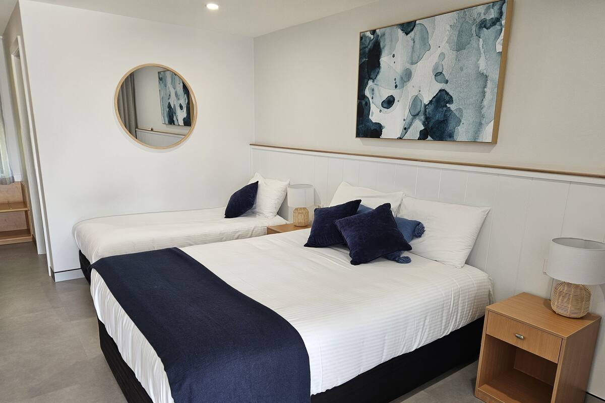 Quarto de hotel com duas camas, um espelho redondo, arte abstrata, uma mesa de cabeceira e um abajur.