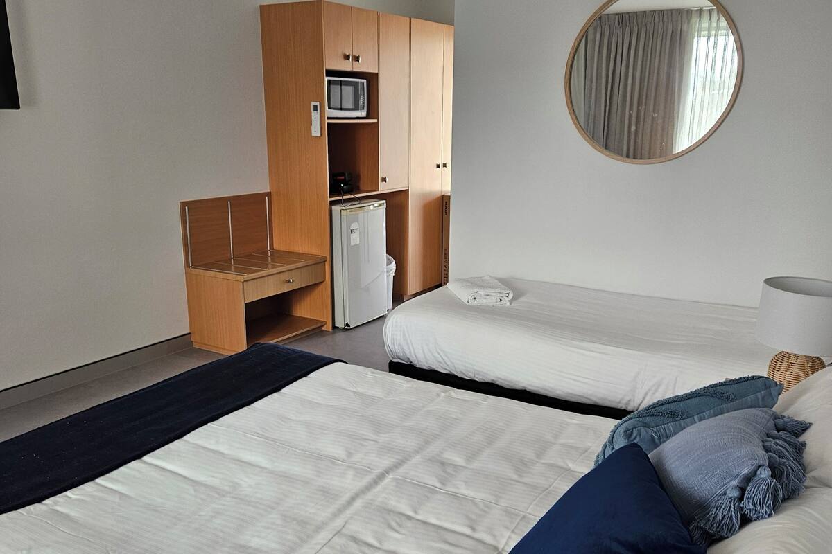 Quarto de hotel com duas camas, uma kitchenette pequena, um espelho redondo e um armário de madeira.