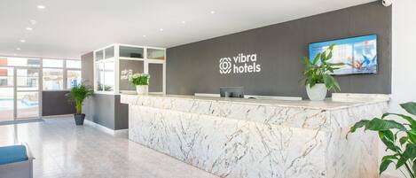 Uma mesa de recepção com tampo de mármore, uma tela grande exibindo uma vista urbana e plantas em vasos.