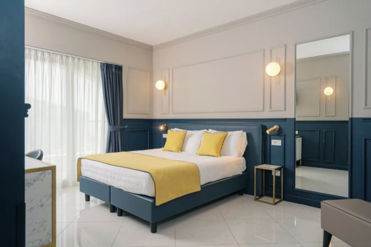 Quarto com cama, mesas de cabeceira, espelho e janela com cortinas.