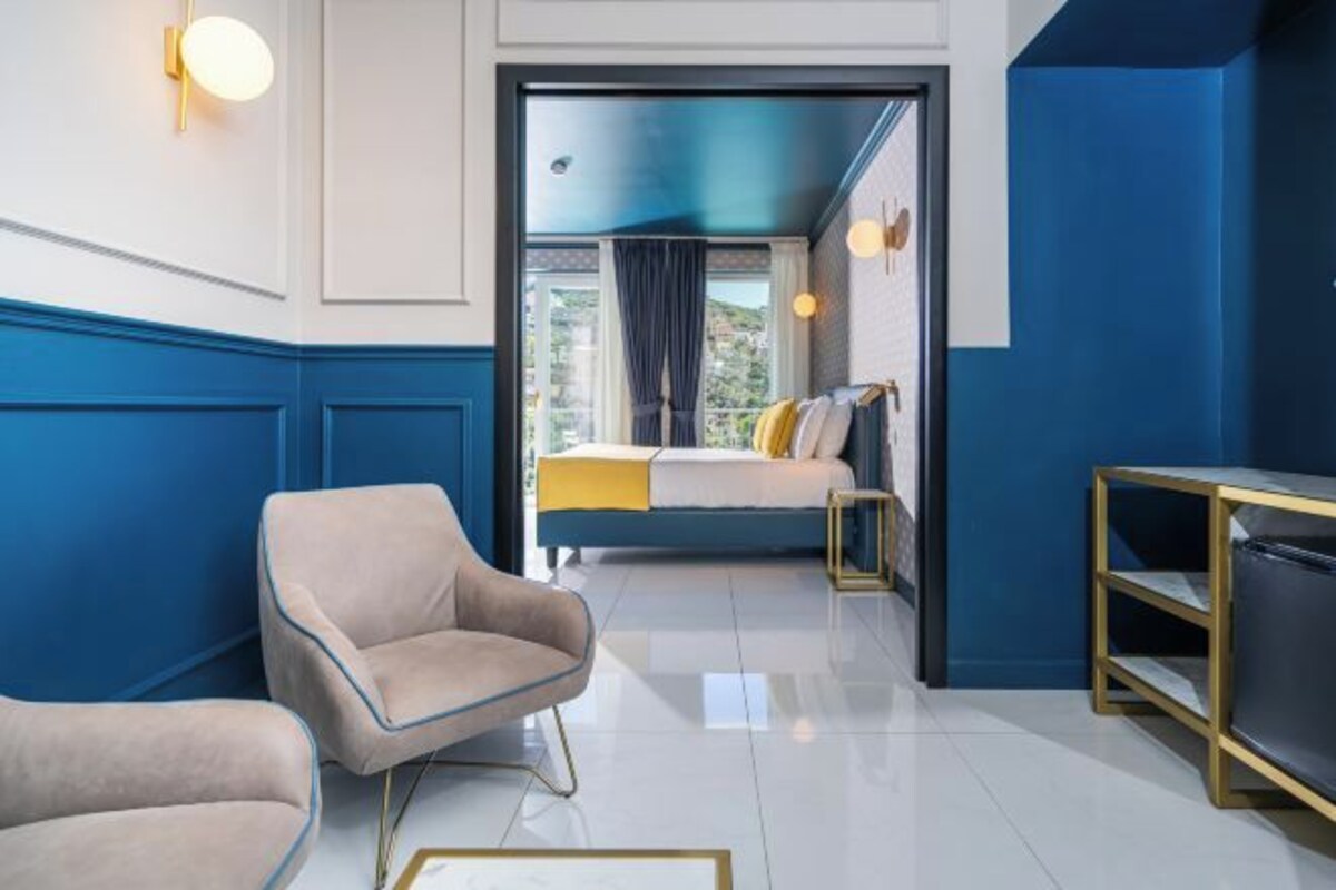 Quarto de hotel moderno com esquema de cores azul e branco, uma cama com cabeceira amarela e azul, uma poltrona bege e uma mesinha de cabeceira com moldura dourada.