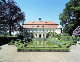 Um edifício amplo com fachada simétrica, um pátio central com fonte e jardins bem cuidados.