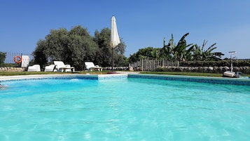 Uma piscina com água azul cristalina, rodeada por espreguiçadeiras brancas e um muro de pedra.