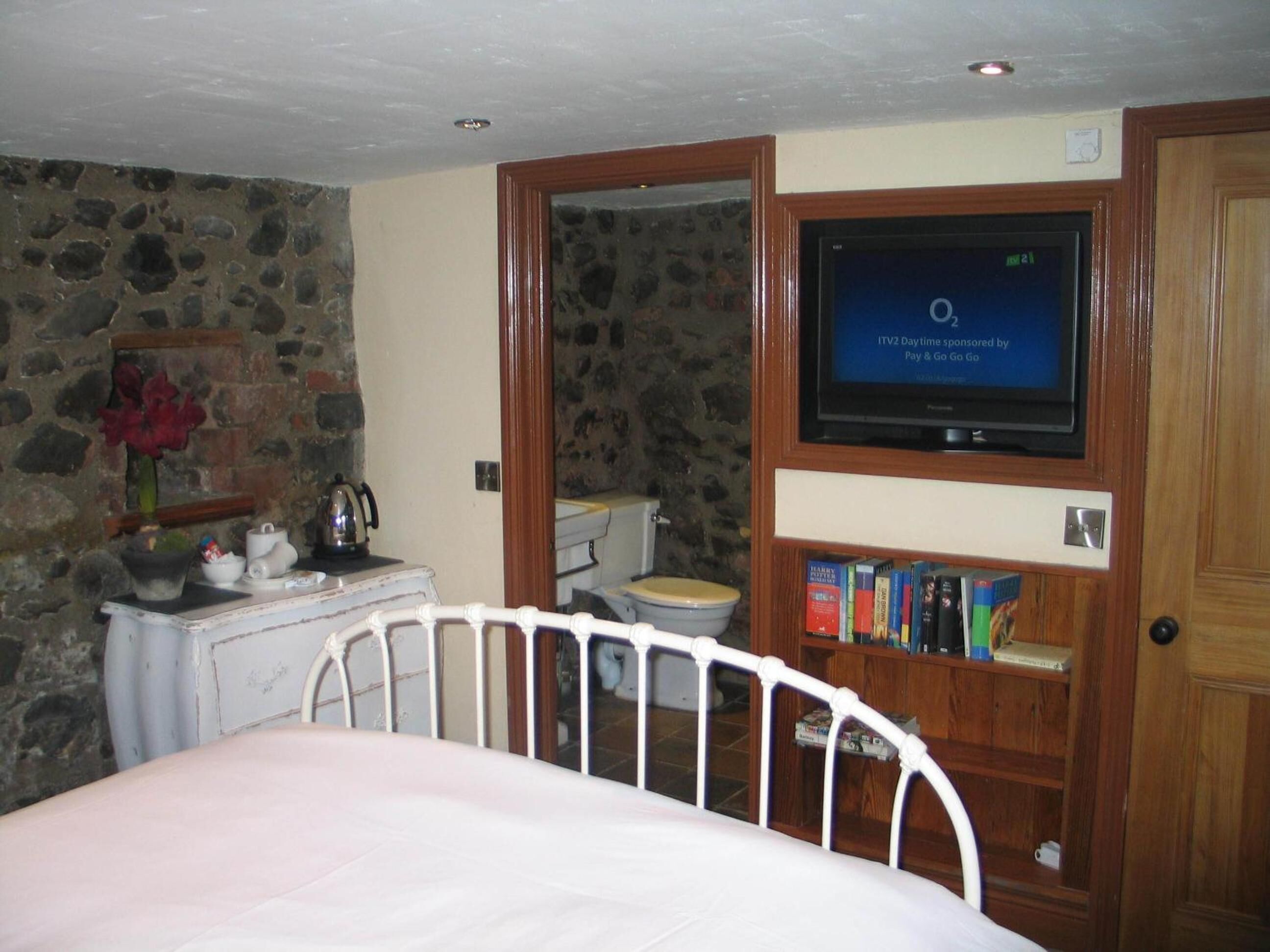 Quarto com cama, televisão na parede, mesa de cabeceira com livros e parede de pedra.