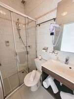 Casa de banho com cabine de duche em vidro, sanita, lavatório com espelho e secador de cabelo fixado na parede.