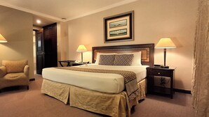 Quarto de hotel com cama, duas mesas de cabeceira, uma cadeira, escrivaninha e abajur.